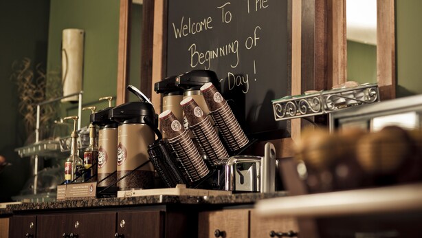 Uma estação de café com uma lousa exibindo a mensagem 'Bem-vindos ao Início do Dia!' e diversos equipamentos para preparo de café.