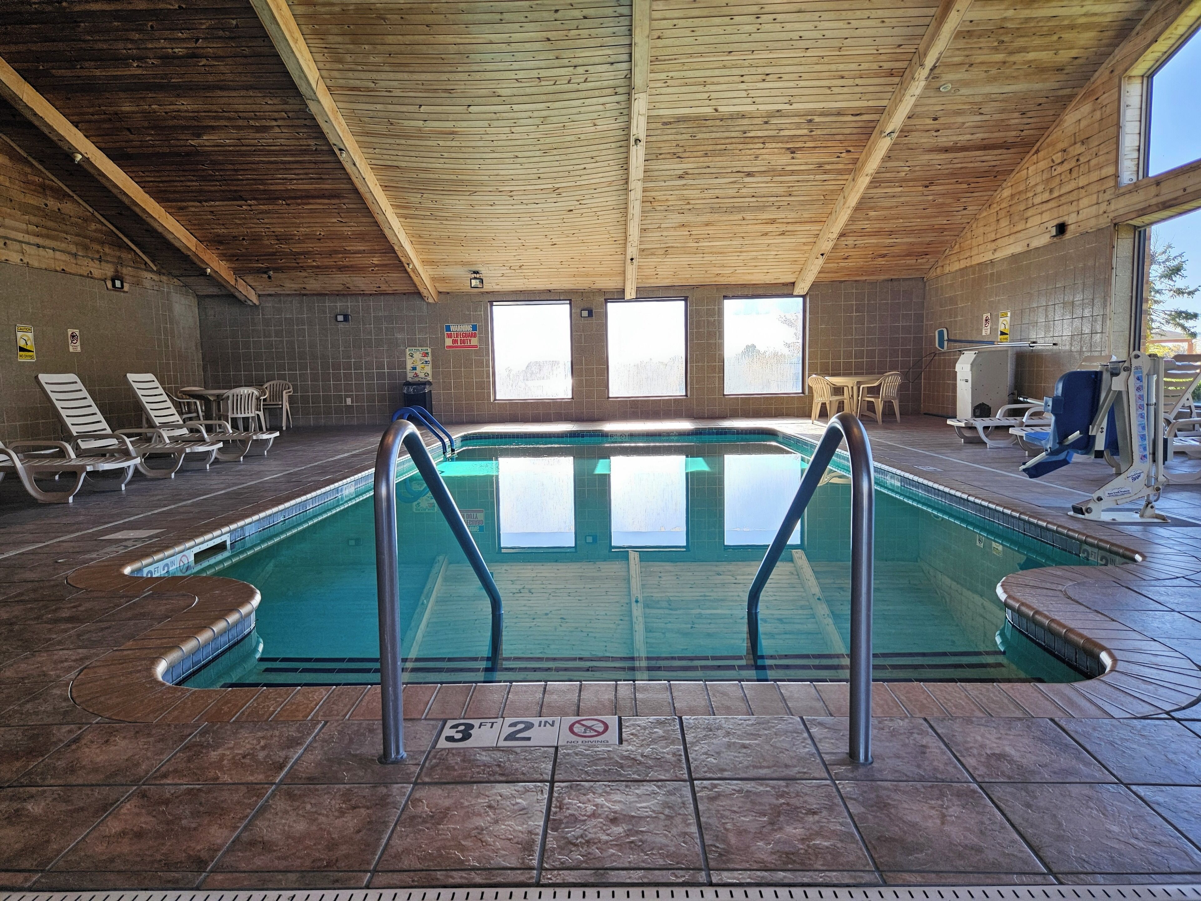 Piscina interna com teto de madeira, paredes azulejadas e ampla vista do céu através de grandes janelas.