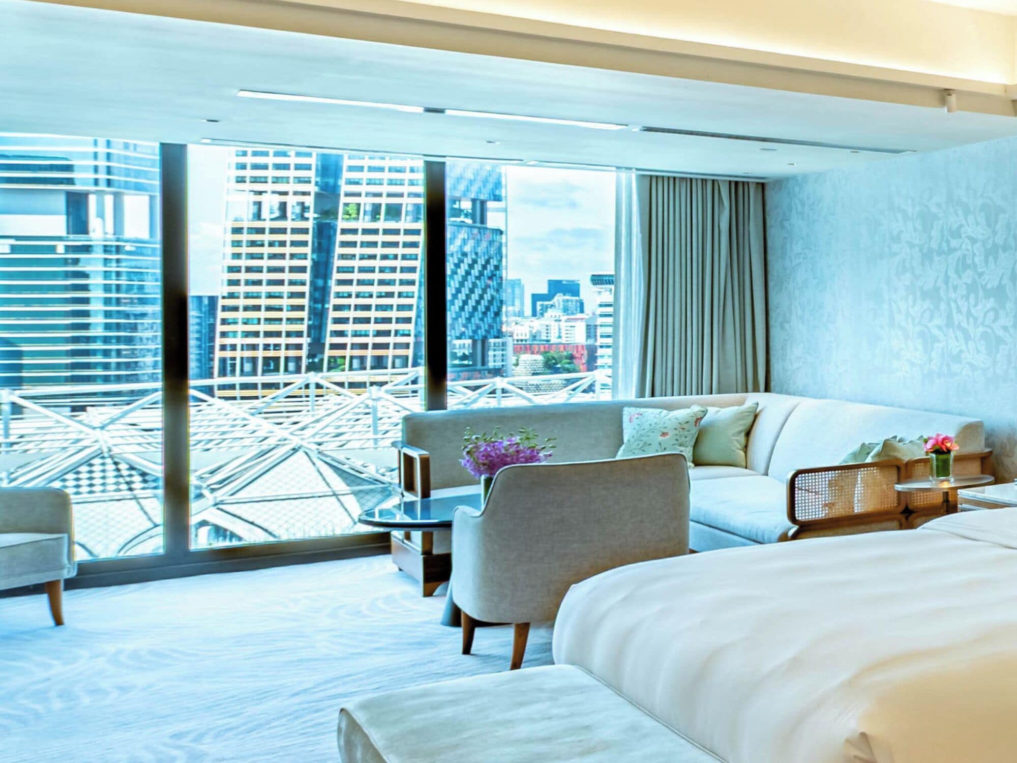 Quarto de hotel moderno com uma cama grande, um sofá, uma poltrona e uma pequena mesa. Há uma janela ampla com vista para a paisagem urbana.