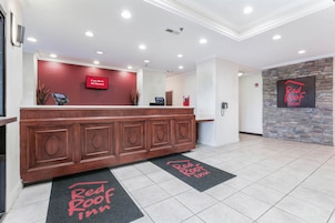 Uma área de recepção de hotel com um balcão de madeira, uma parede vermelha com o aviso 'Wi-Fi Gratuito em Todos os Quartos', uma parede de pedra com o anúncio 'Red Roof Inn' e um tapete de entrada preto e vermelho.