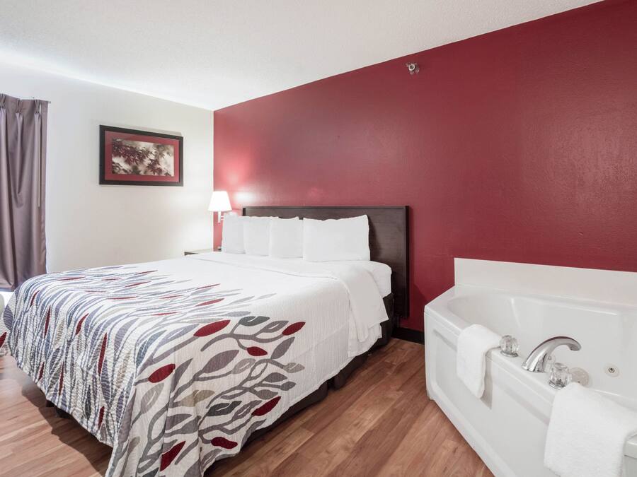 Quarto de hotel com cama, luminária de cabeceira, janela grande com cortinas, quadro na parede e banheiro privativo com banheira.