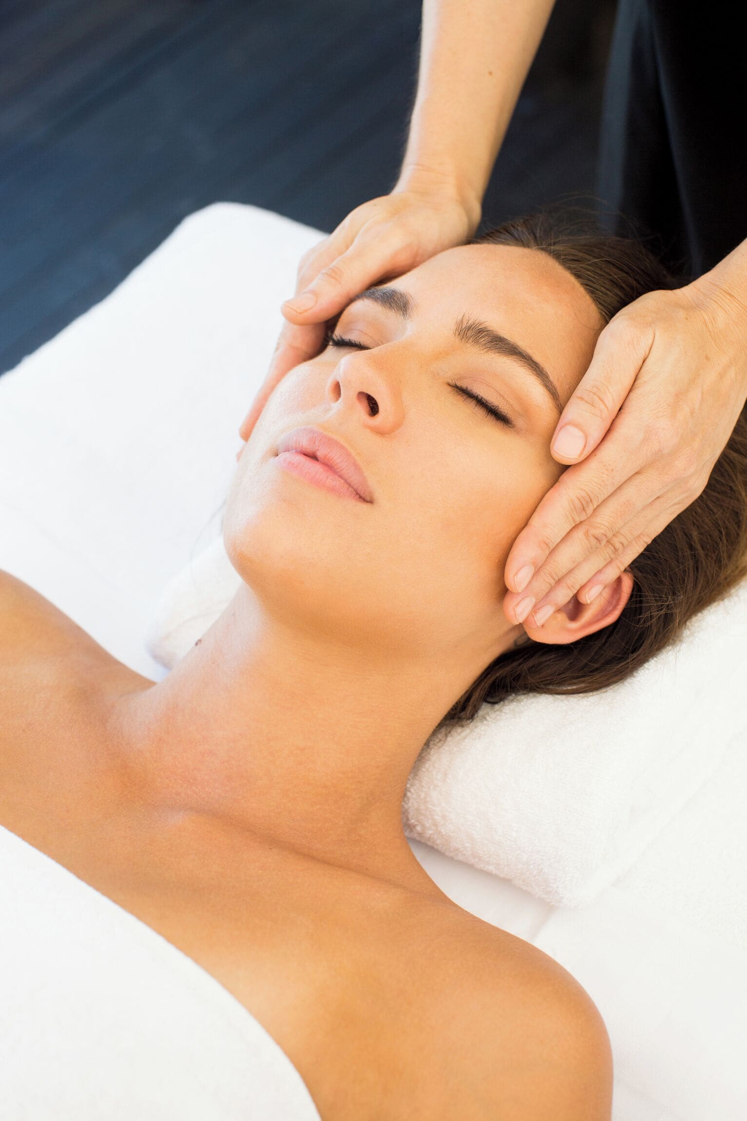 Uma pessoa deitada em uma superfície branca com uma toalha, recebendo massagem na cabeça.