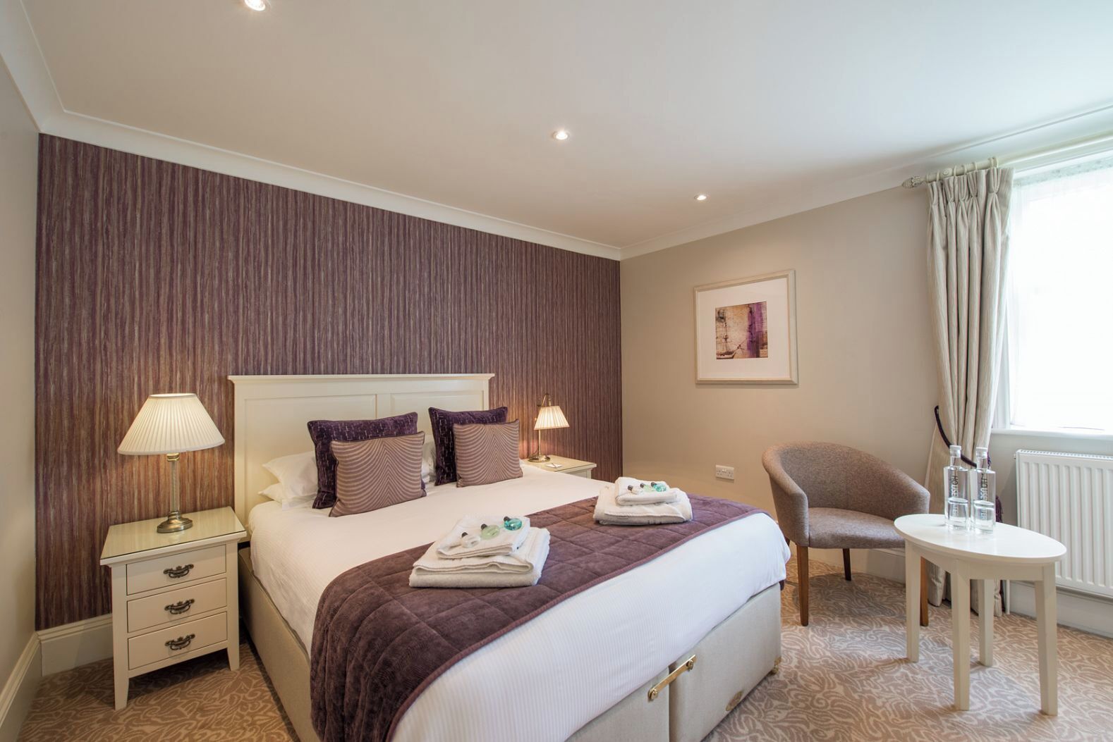Quarto de hotel com uma cama grande, duas mesas de cabeceira, uma cadeira e uma pequena mesa com garrafas.