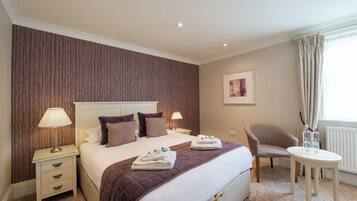 Quarto de hotel com uma cama grande, duas mesas de cabeceira, uma cadeira e uma pequena mesa com garrafas.