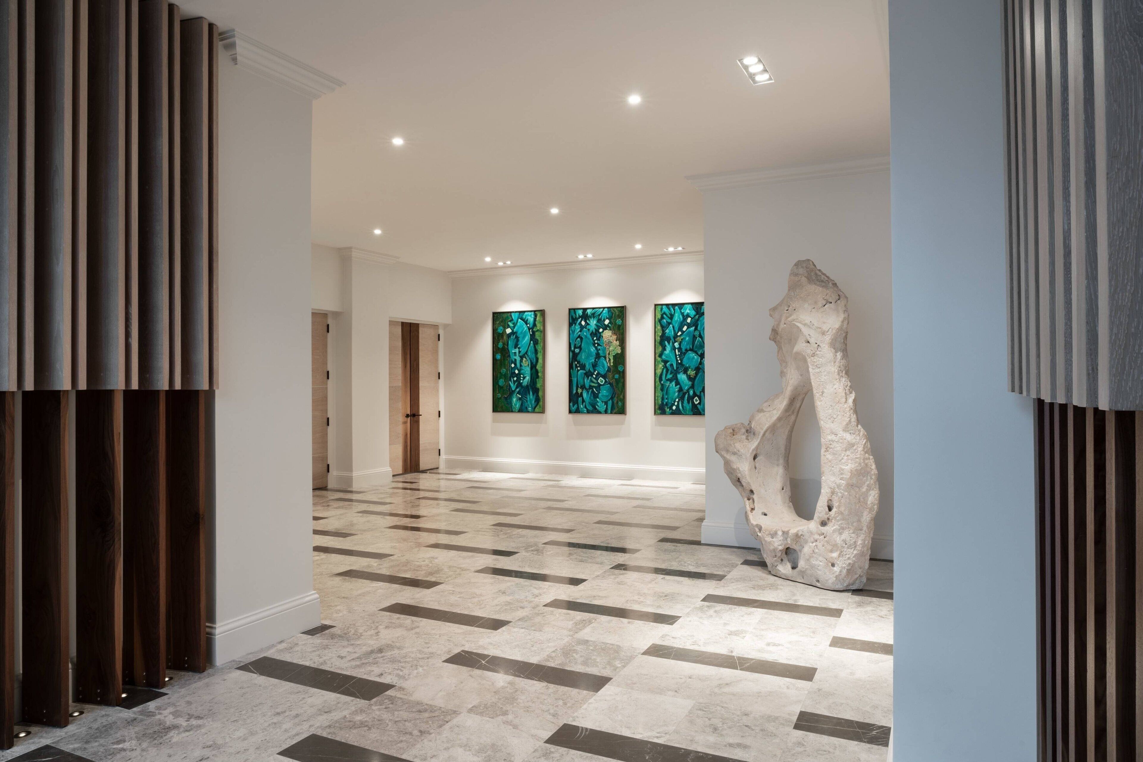 Um corredor espaçoso com piso xadrez, uma grande escultura abstrata e três pinturas verdes na parede.