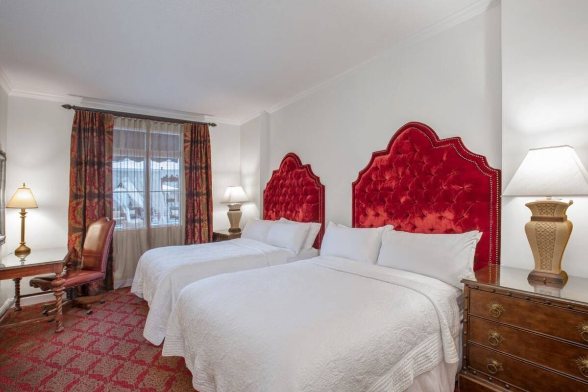 Quarto de hotel com duas camas, uma escrivaninha com cadeira, uma luminária e um armário.
