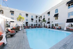 Uma área de piscina com espreguiçadeiras e guarda-sóis, uma piscina azul límpida e um edifício branco com várias janelas e varandas.