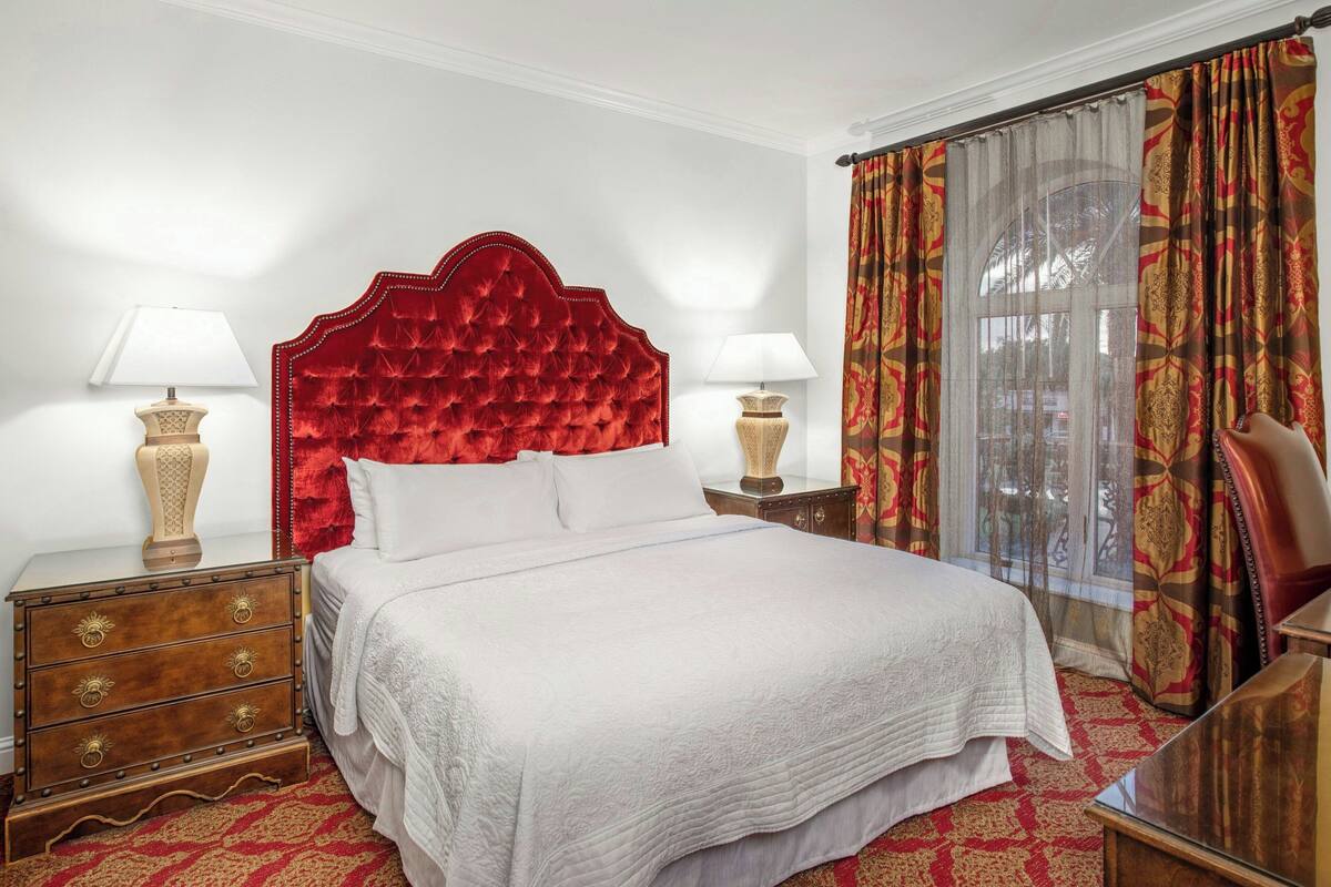 Quarto com cabeceira estofada vermelha, roupa de cama branca, duas mesas de cabeceira com abajures e uma janela grande com cortinas estampadas.