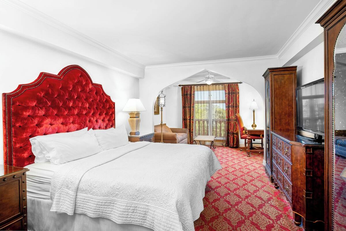 Quarto de hotel com uma cama grande, penteadeira de madeira, televisão e piso coberto por carpete vermelho.