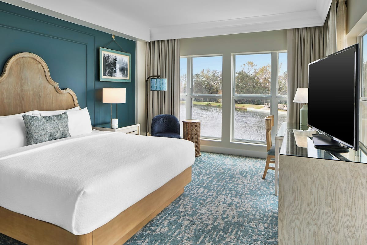 Quarto de hotel com uma cama grande, televisão, escrivaninha e vista para a água através de amplas janelas.