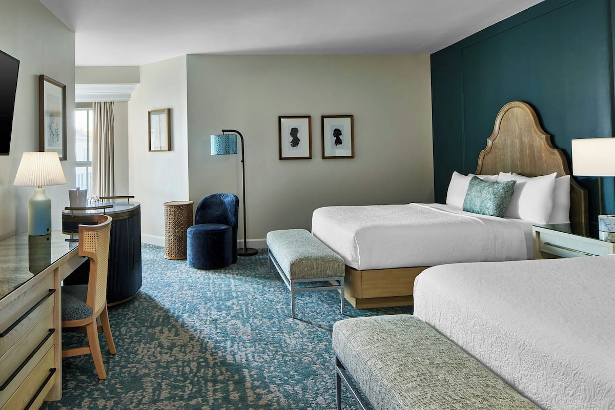 Quarto de hotel com duas camas, uma escrivaninha, uma cadeira, um abajur e obras de arte emolduradas na parede.