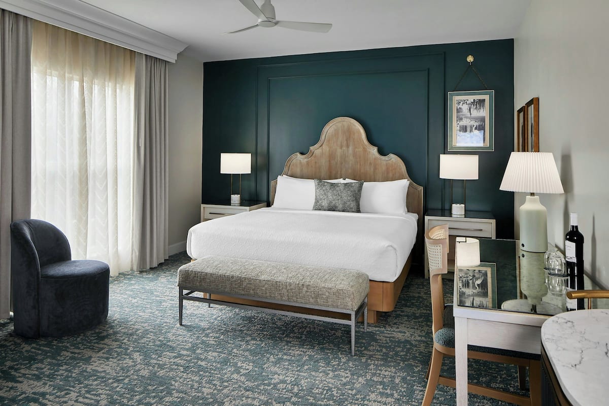 Quarto de hotel com uma cama grande, uma mesa com cadeira, uma mesinha e um ventilador de teto.
