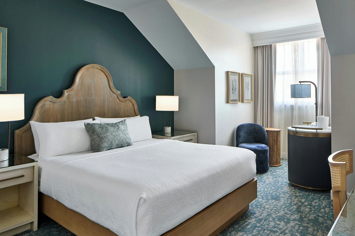 Quarto de hotel com uma cama grande, duas mesas de cabeceira, uma cadeira, um abajur e uma janela com cortinas.