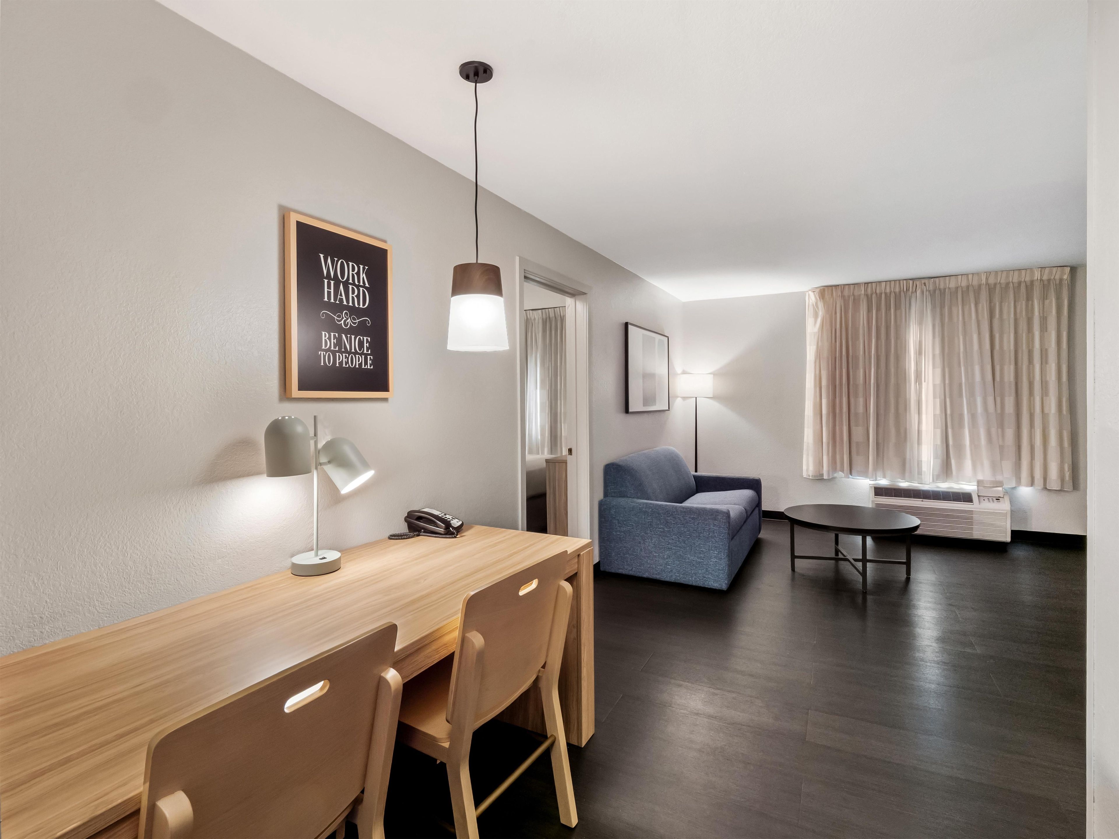 Quarto de hotel com escrivaninha de madeira, duas cadeiras, um sofá, uma mesinha, um abajur, ar-condicionado e uma citação motivacional emoldurada na parede.