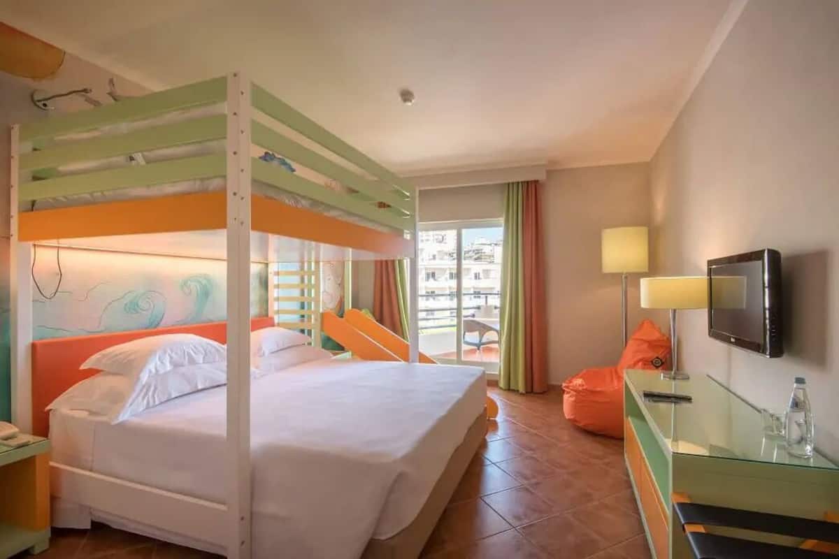 Quarto de hotel com beliche, escrivaninha de vidro, televisão e varanda com vista para edifícios.