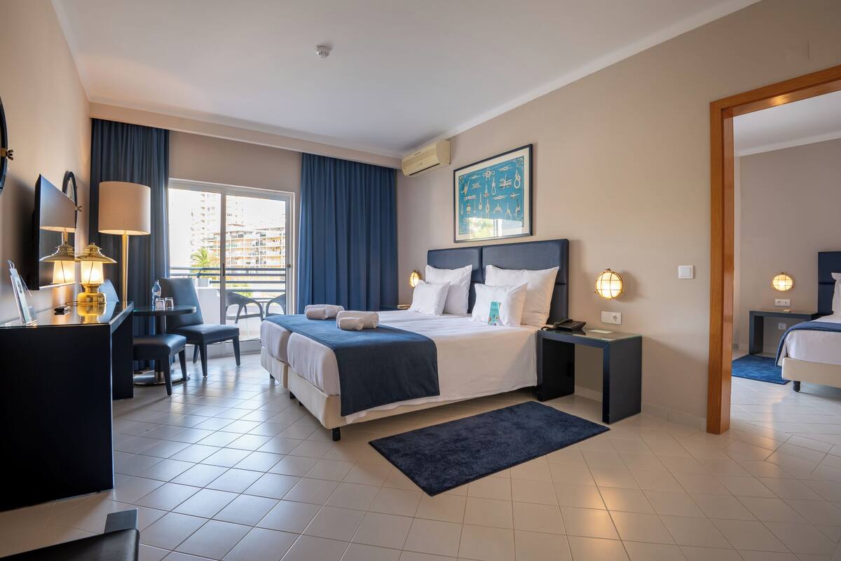 Quarto de hotel com uma cama grande, duas mesas de cabeceira, uma escrivaninha com cadeira, uma luminária de mesa e uma janela ampla com cortinas.