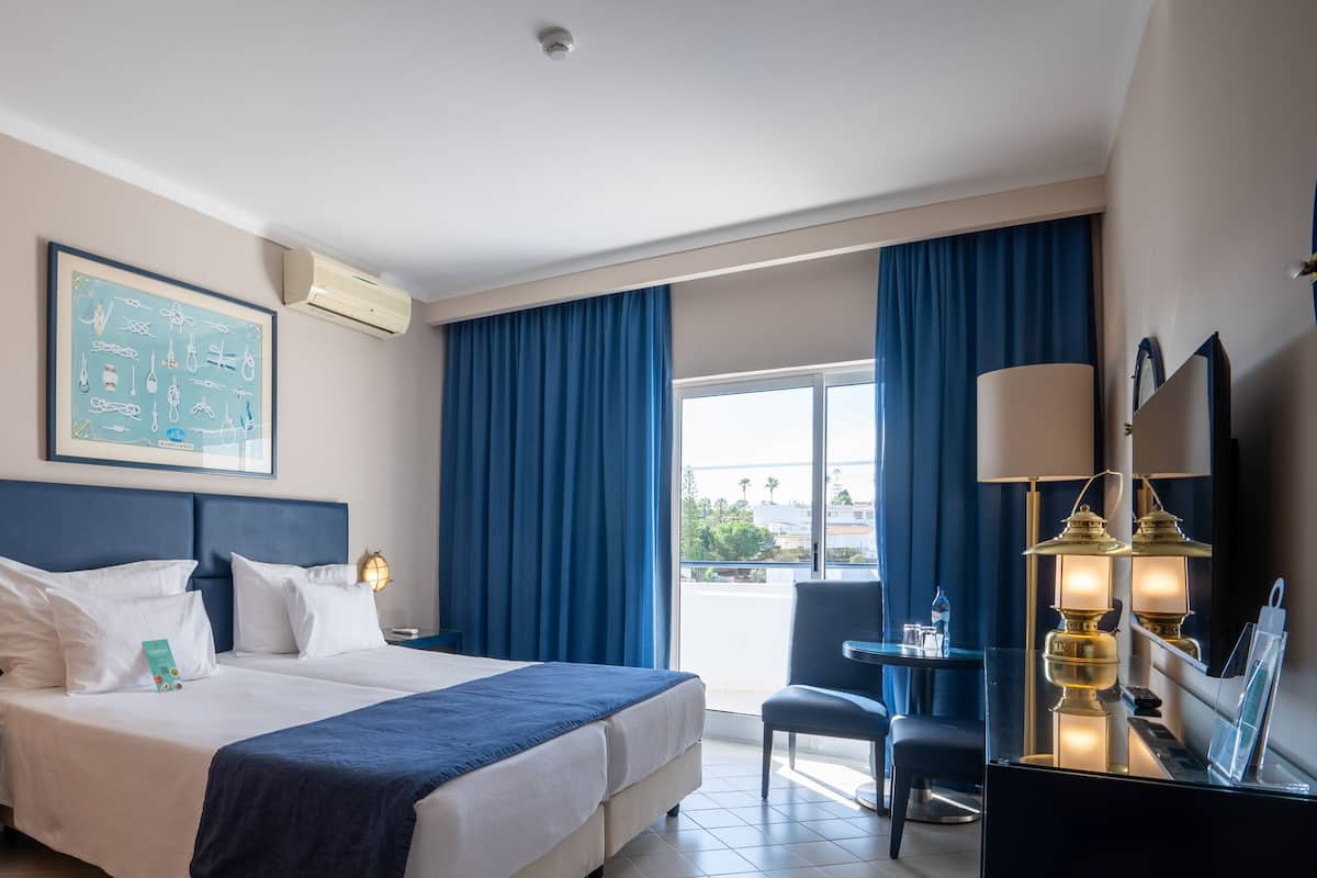 Quarto de hotel com cama grande, escrivaninha, cadeira, luminária e janela com cortinas.