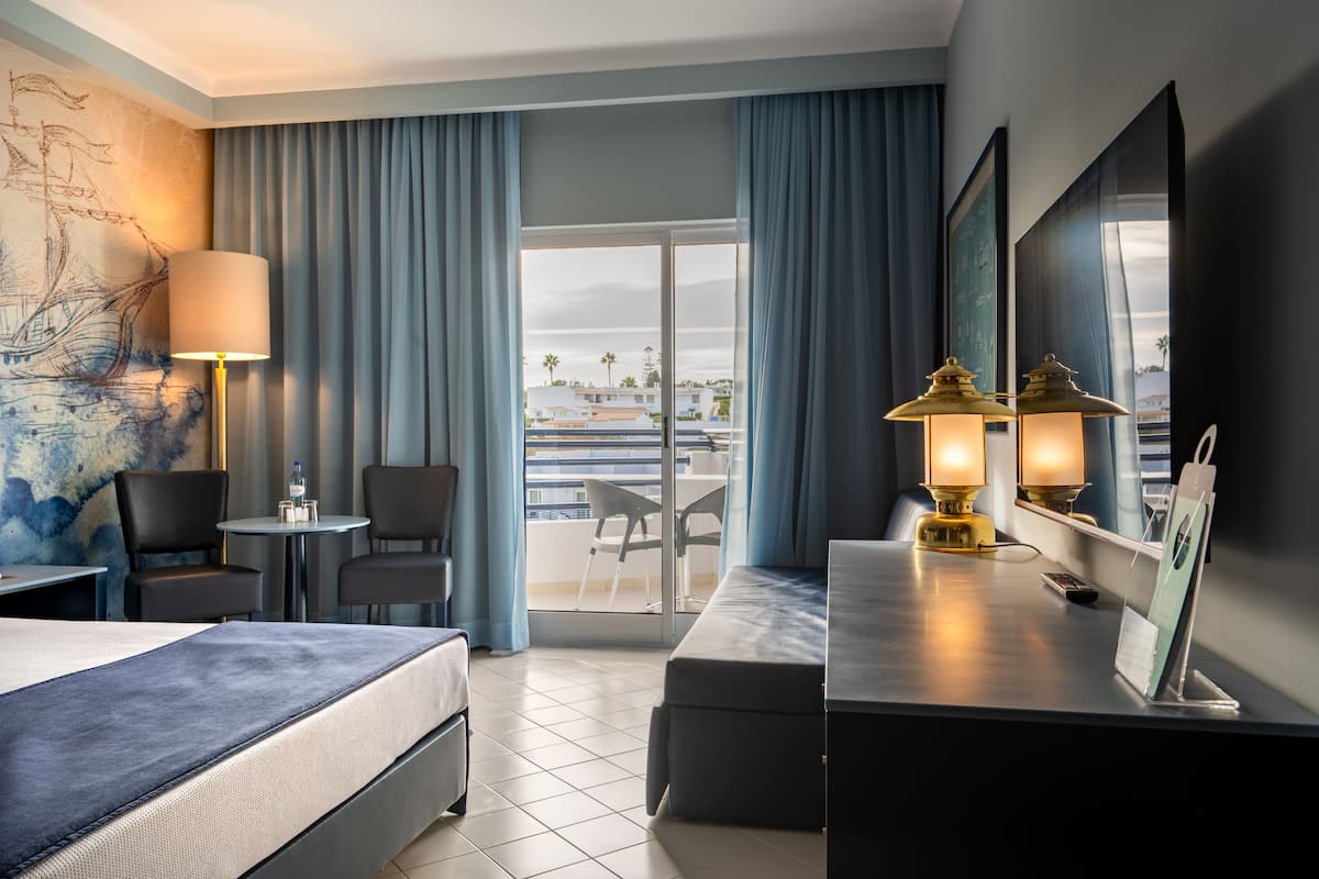 Quarto de hotel com cama, duas cadeiras, uma mesinha, luminária, televisão e varanda com vista para a cidade.