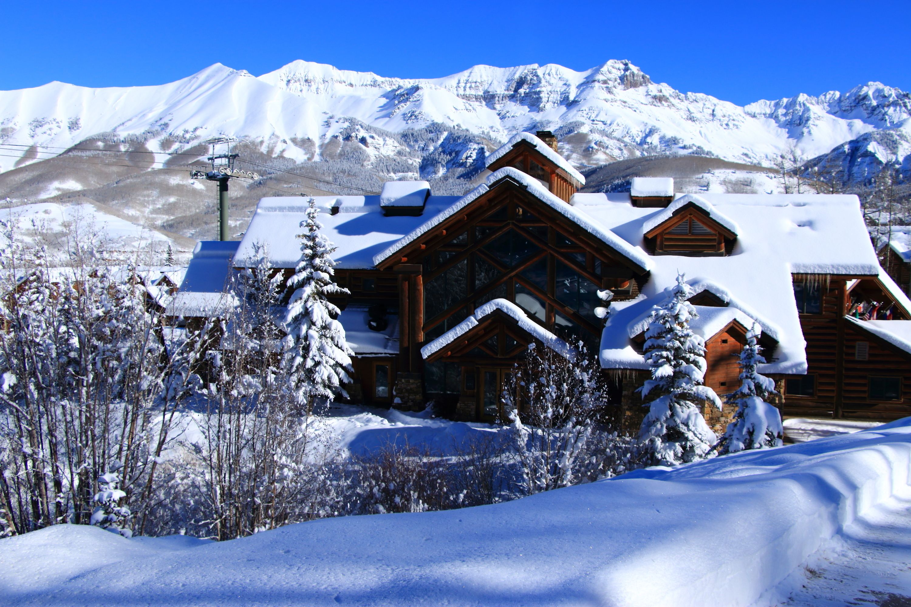 Uma cabana de tronco com telhado coberto de neve e janelas grandes, rodeada por árvores carregadas de neve e montanhas ao fundo.