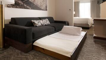 Quarto de hotel com sofá-cama, cama de casal, televisão de tela plana e uma janela grande com cortinas.