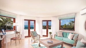 Uma sala de estar espaçosa com vista para o oceano, uma escrivaninha branca com abajur, um sofá verde e cadeiras de vime.