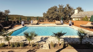 Uma piscina retangular com um deck de pedra, rodeada por espreguiçadeiras e guarda-sóis, com árvores e colinas ao fundo.