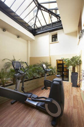 Ginásio interior com equipamentos de exercício, piso de madeira e plantas em vasos.