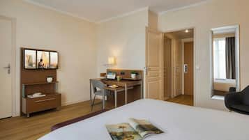 Quarto de hotel com cama, escrivaninha, cadeira, estante de livros e janela com cortinas.