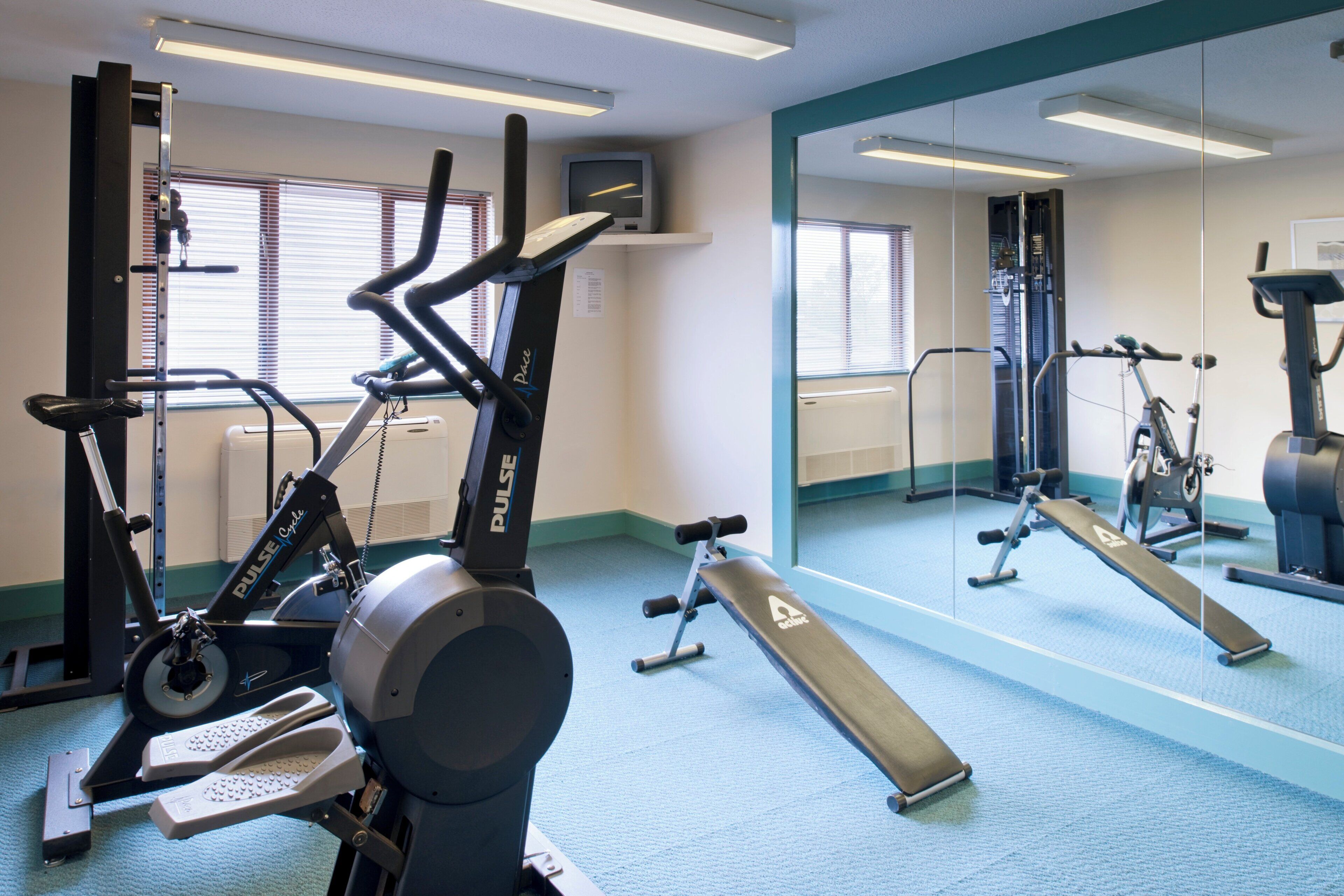 Academia com equipamentos de exercícios, parede espelhada e piso coberto por carpete azul.