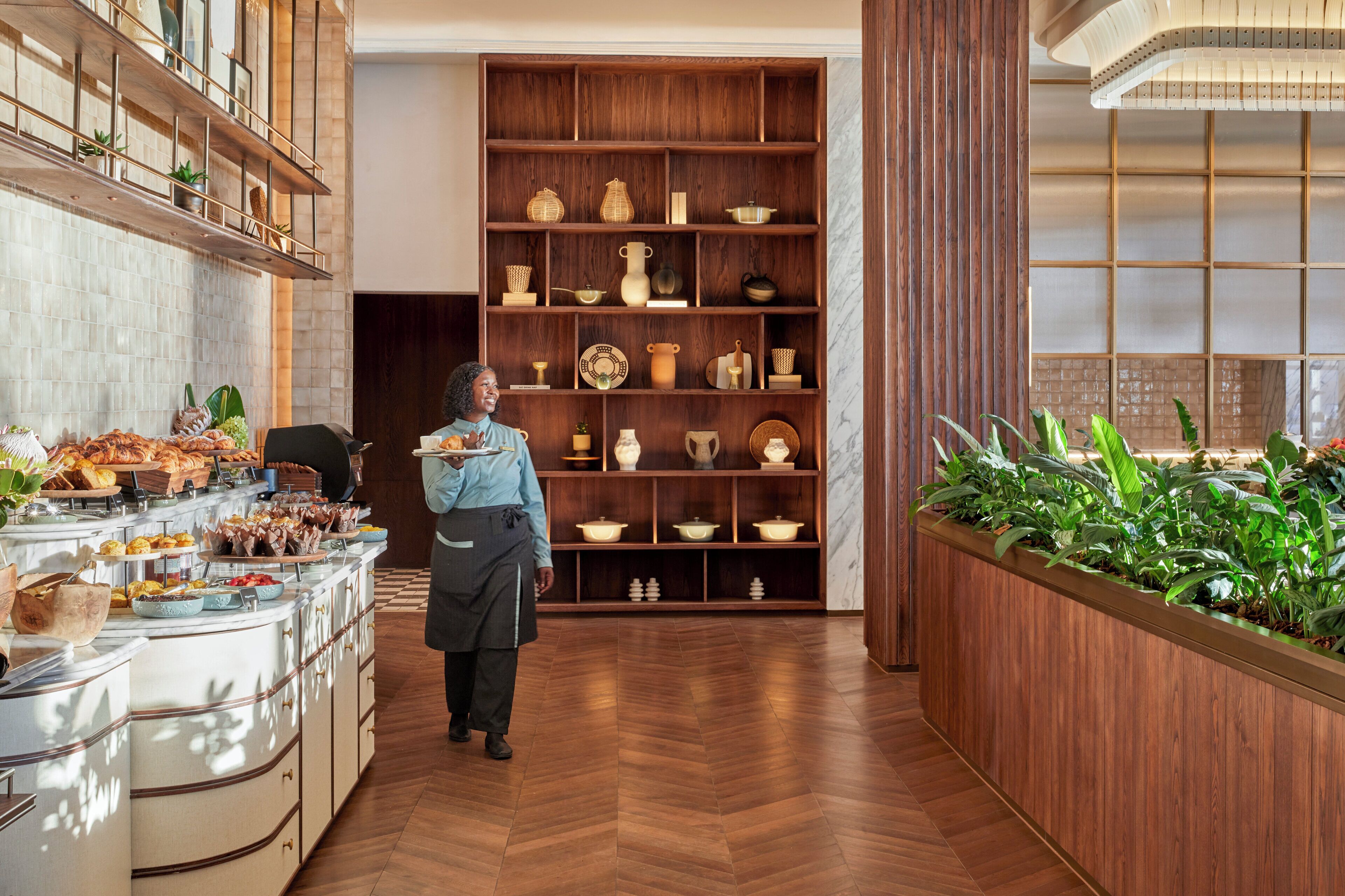 Um saguão de hotel com área de buffet, uma grande estante de madeira com objetos decorativos e um vaso com plantas verdes.