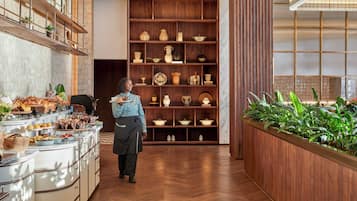 Um saguão de hotel com área de buffet, uma grande estante de madeira com objetos decorativos e um vaso com plantas verdes.