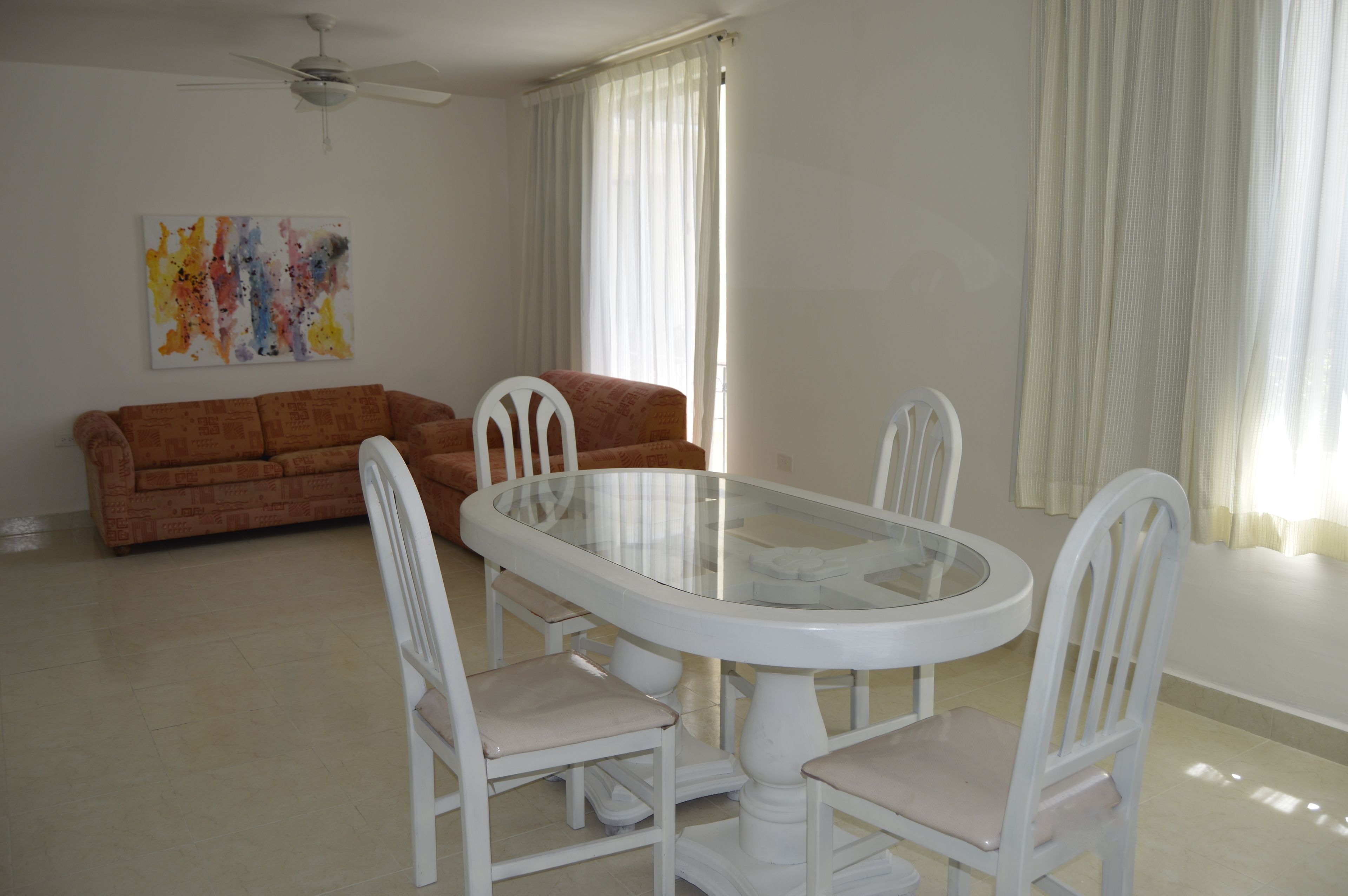 Uma área de refeições com mesa de vidro e cadeiras brancas, um sofá marrom e um ventilador de teto.