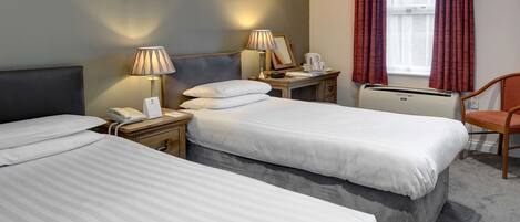 Quarto de hotel com duas camas, luminárias de cabeceira, uma escrivaninha com telefone, uma cadeira e uma janela com cortinas.