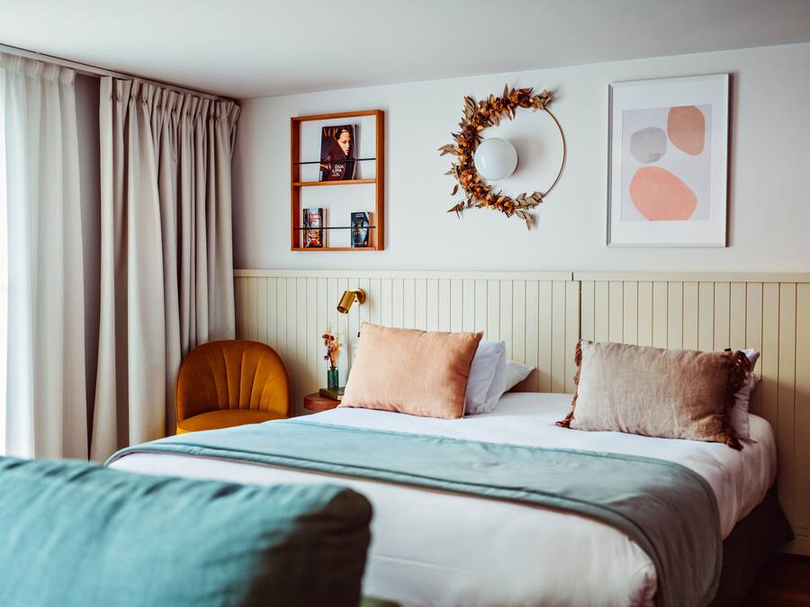 Quarto bem organizado com cama, mesa de cabeceira, cadeira e prateleira fixada na parede com livros e uma foto emoldurada.
