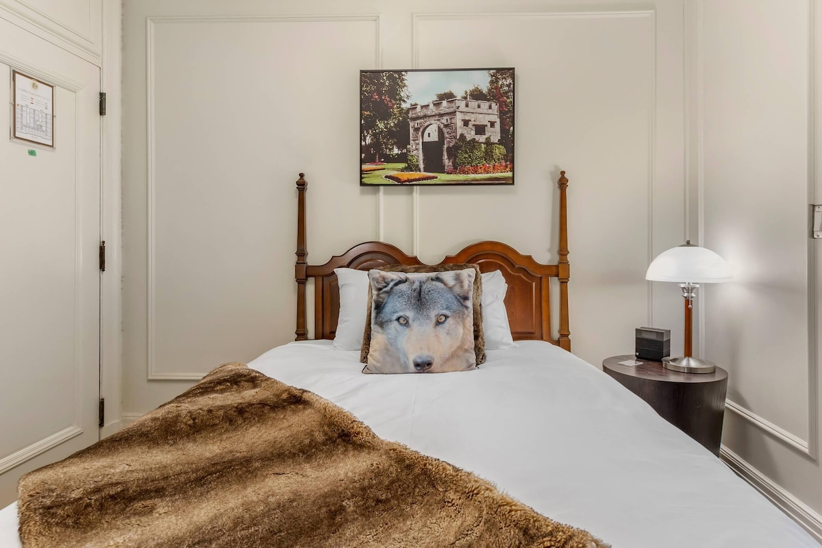 Quarto de hotel com cama, mesa de cabeceira com abajur, quadro pendurado na parede e porta com aviso.