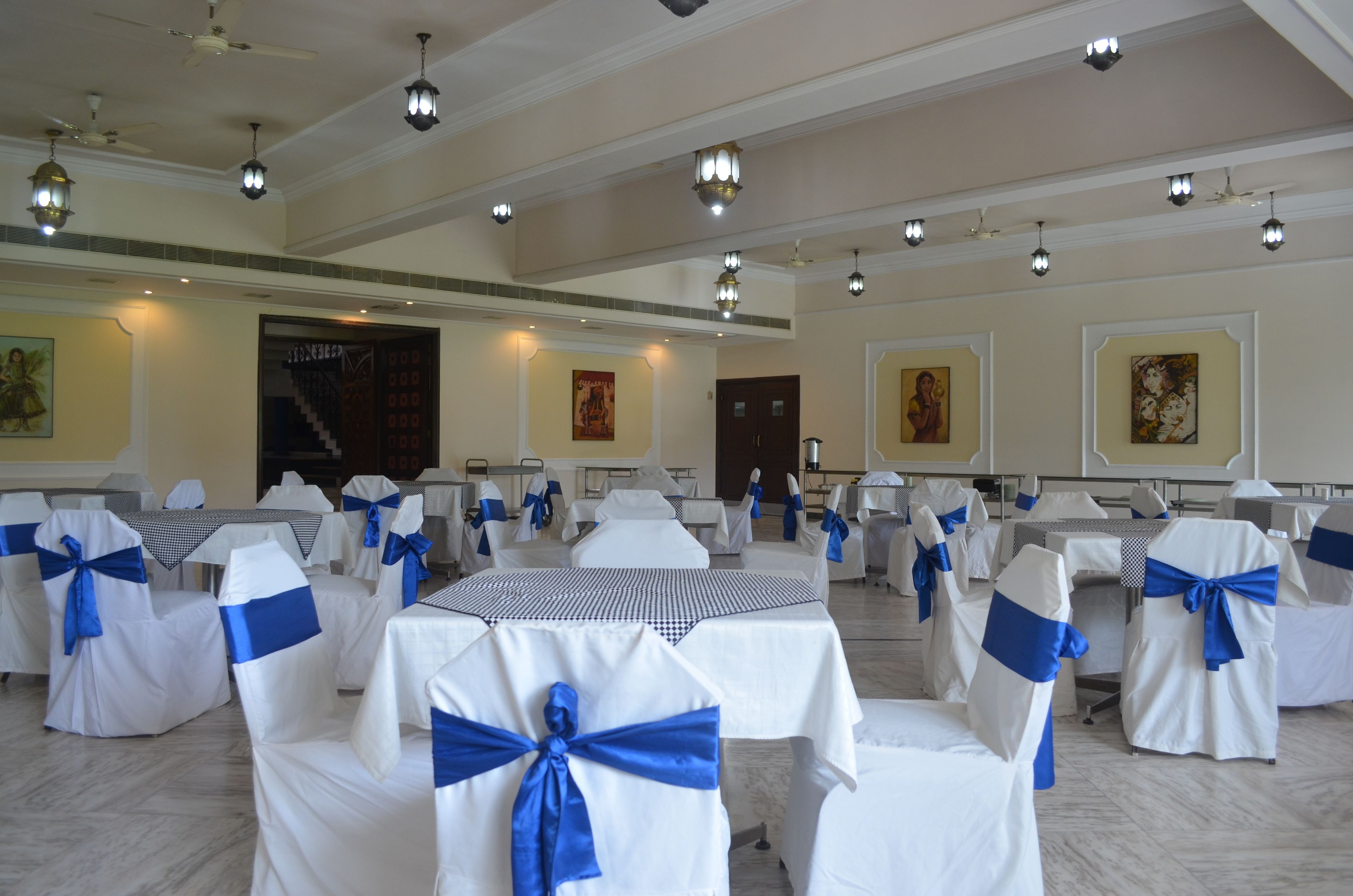 Uma sala de banquetes com mesas e cadeiras brancas, fitas azuis e lustres.