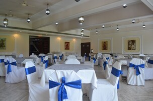 Uma sala de banquetes com mesas e cadeiras brancas, fitas azuis e lustres.