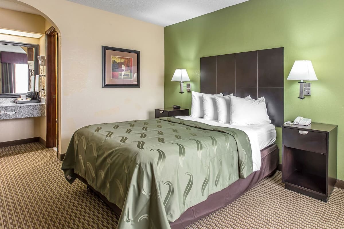 Quarto de hotel com cama, duas luminárias de cabeceira, mesa de cabeceira com telefone, quadro na parede e banheiro com pia e espelho.