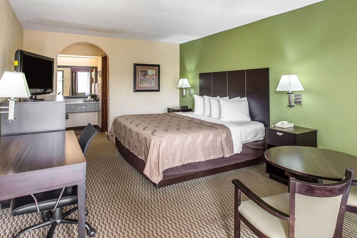 Quarto de hotel com cama, escrivaninha, cadeira e televisão.
