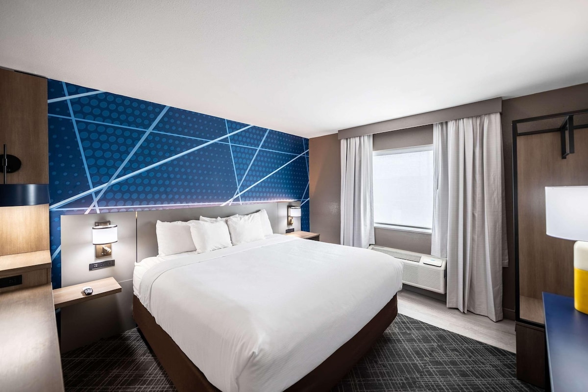Quarto de hotel moderno com uma cama grande, mesa de cabeceira, luminária na parede, janela com cortinas e vista para o exterior.