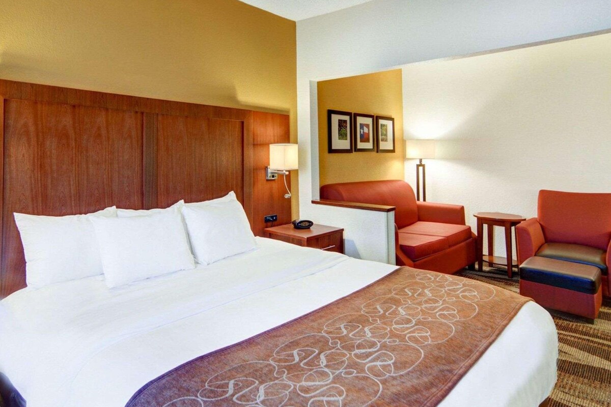 Quarto de hotel com uma cama grande, cabeceira de madeira, escrivaninha com luminária, poltrona vermelha e uma pequena mesa.