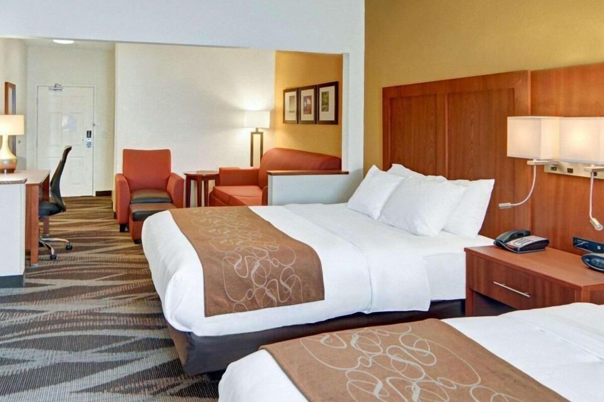 Quarto de hotel com uma cama grande, uma escrivaninha com cadeira, uma mesinha e uma luminária.