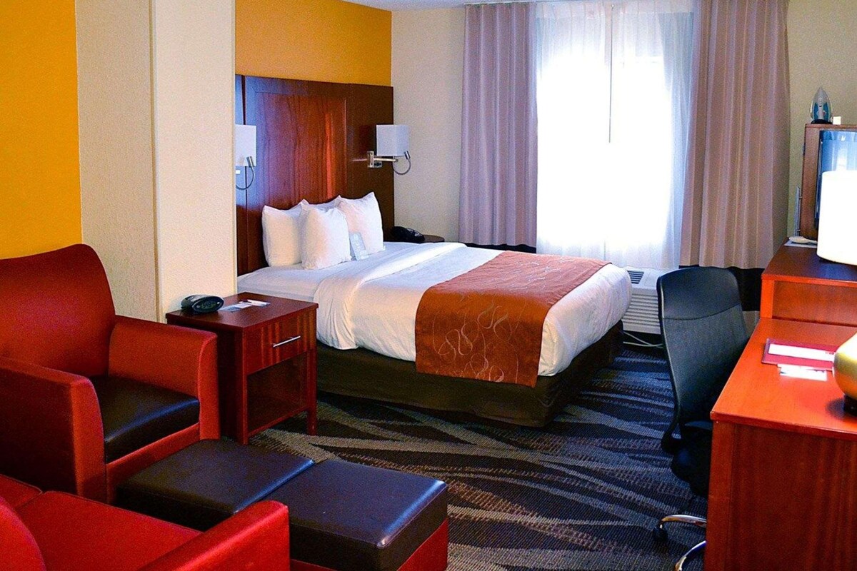 Quarto de hotel com cama, escrivaninha, cadeira, mesa de cabeceira e janela com cortinas.