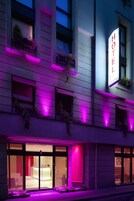 Exterior de hotel com letreiro de neon, varandas com plantas e área de recepção visível através das portas de vidro.