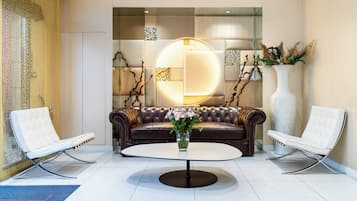 Um lobby moderno com um sofá de couro marrom, duas cadeiras brancas, uma mesa redonda e elementos decorativos, incluindo um vaso grande e arte na parede.