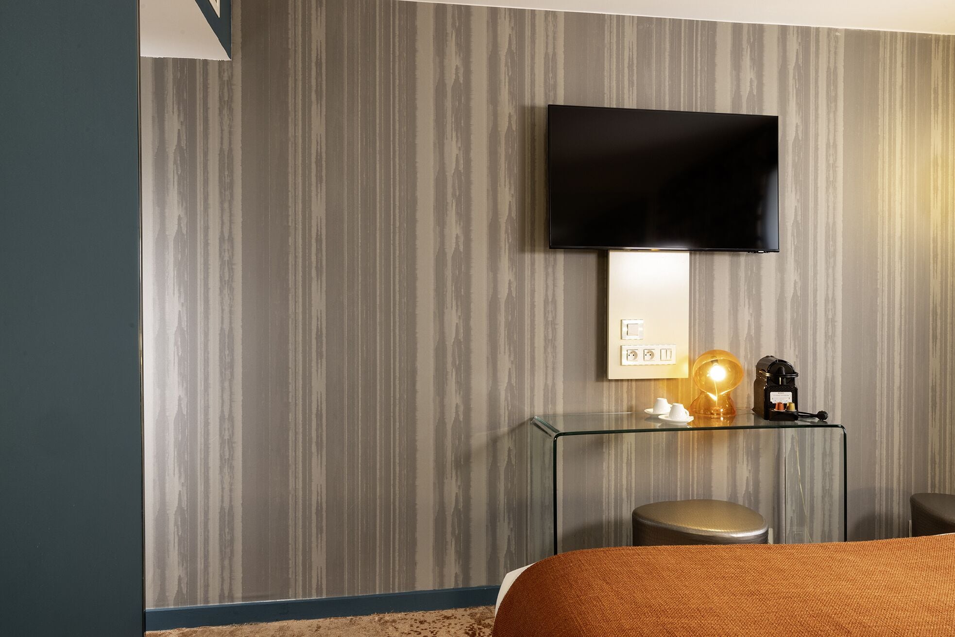 Quarto de hotel com televisão de tela plana na parede, mesa de vidro com cafeteira e cama com cobertor laranja.