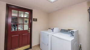 Uma lavanderia com uma porta vermelha, duas máquinas de lavar brancas, uma secadora e um aviso com os horários do centro de atividades físicas.