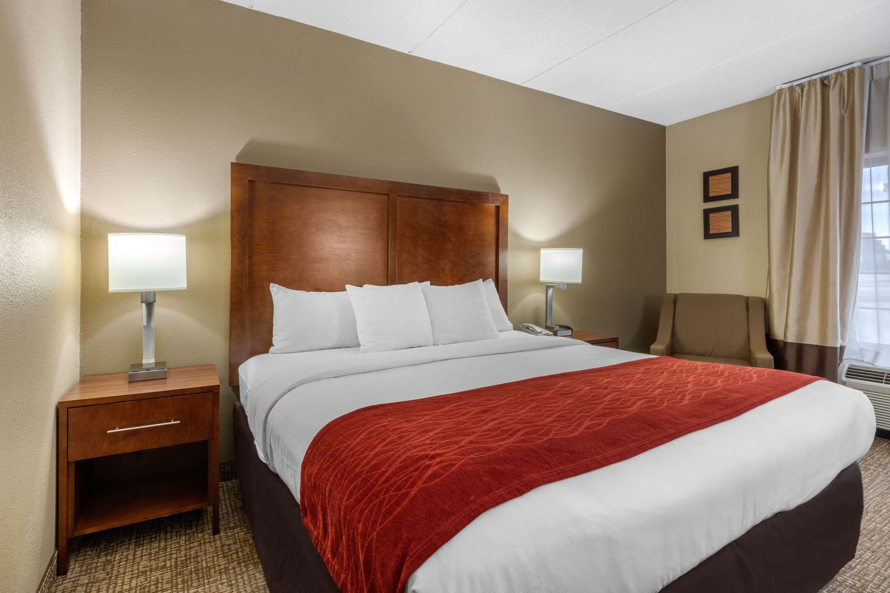 Quarto de hotel com uma cama grande, duas mesas de cabeceira com luminárias, uma cadeira e uma janela com cortinas.