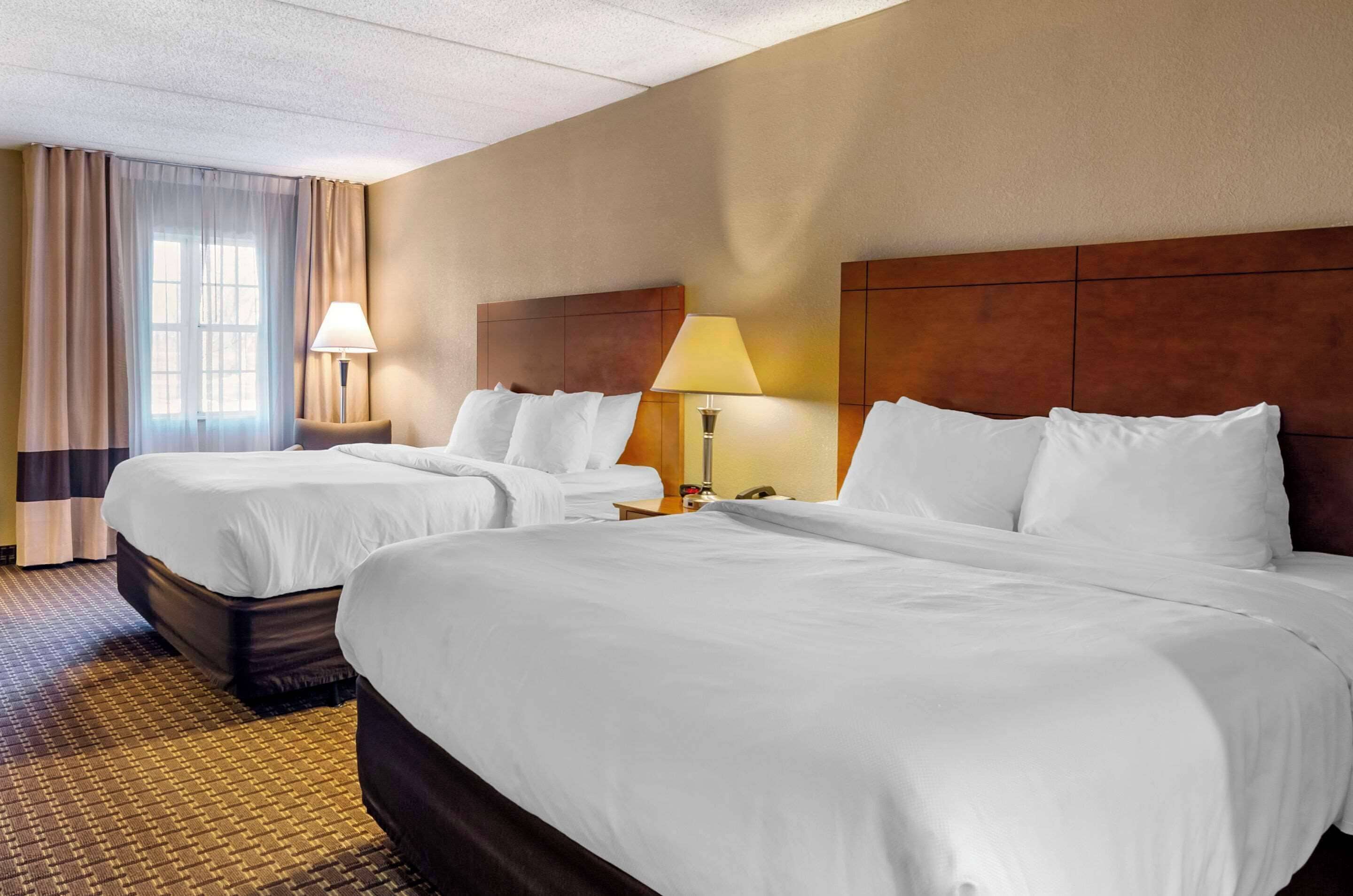 Quarto de hotel com duas camas, uma janela grande com cortinas e uma mesinha de cabeceira com abajur.