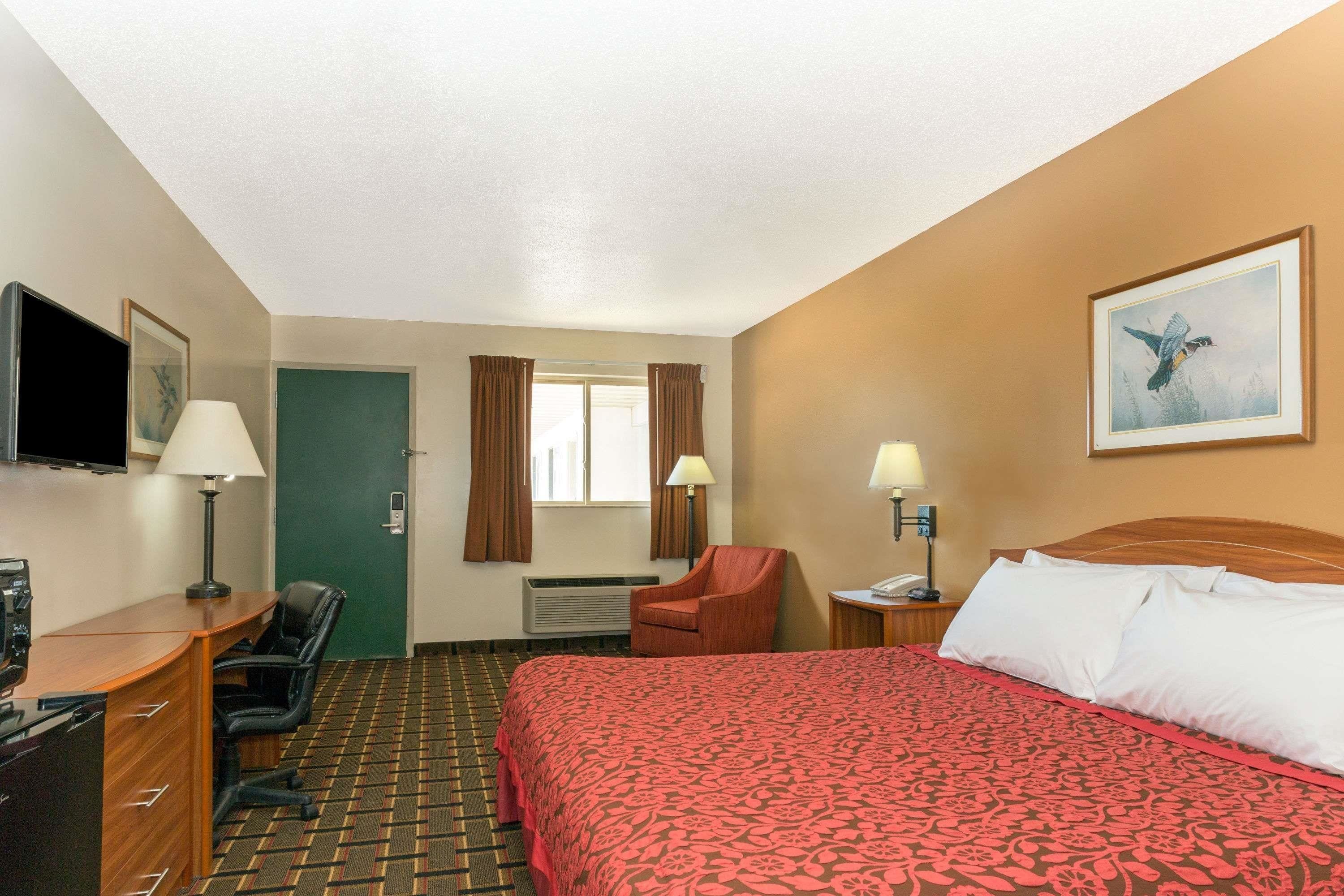 Quarto de hotel com cama, escrivaninha, cadeira, televisão e janela com cortinas.